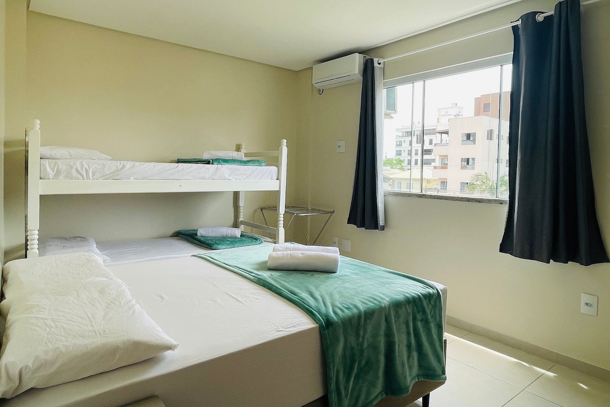 Quarto individual com uma cama de solteiro e um beliche, janela com cortinas e ar condicionado na parede.