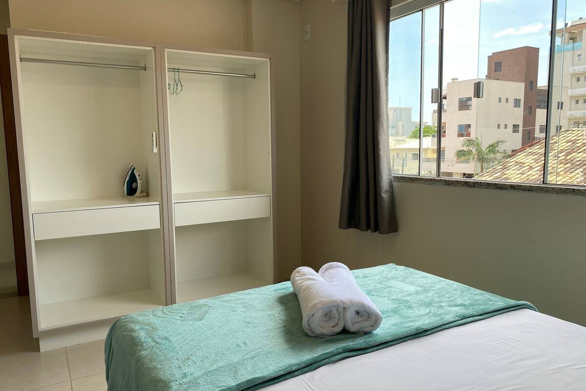 Quarto de hotel com cama, manta verde, dois toalhas, guarda-roupa e janela com vista para edifícios.