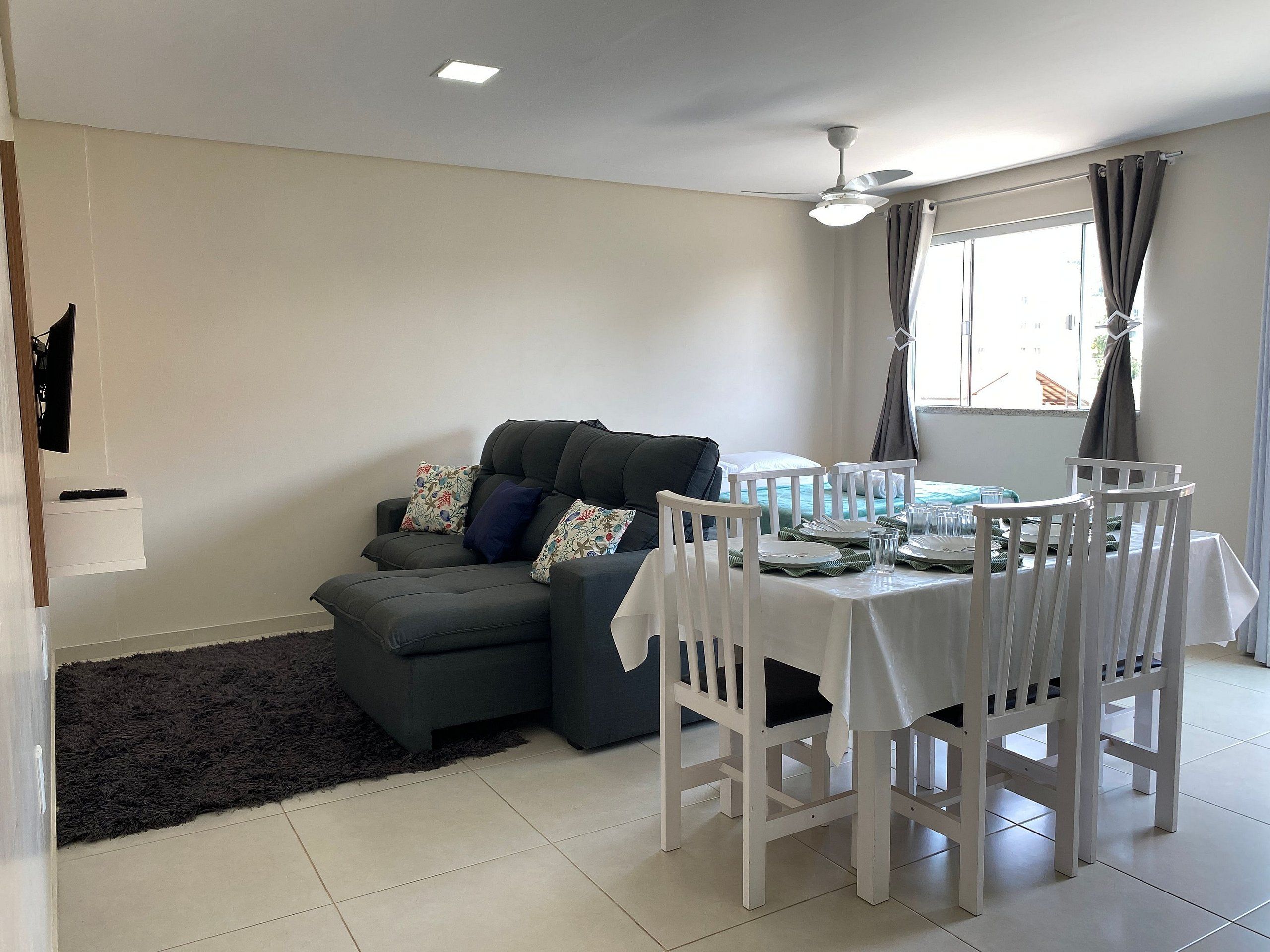 Uma sala de jantar com uma mesa posta para quatro pessoas, um sofá cinza com almofadas estampadas e um ventilador de teto.