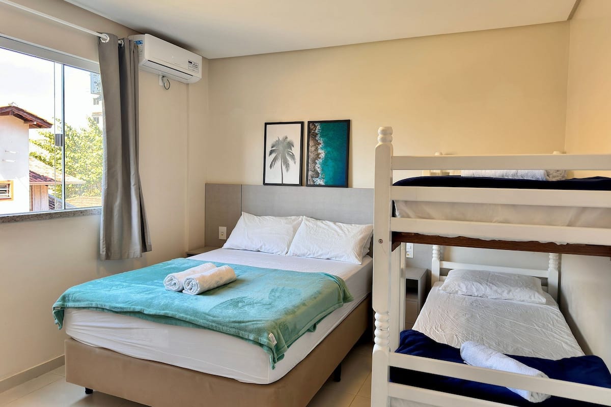 Quarto de hotel com cama, beliche, janela com cortinas, aparelho de ar condicionado e quadro pendurado na parede.