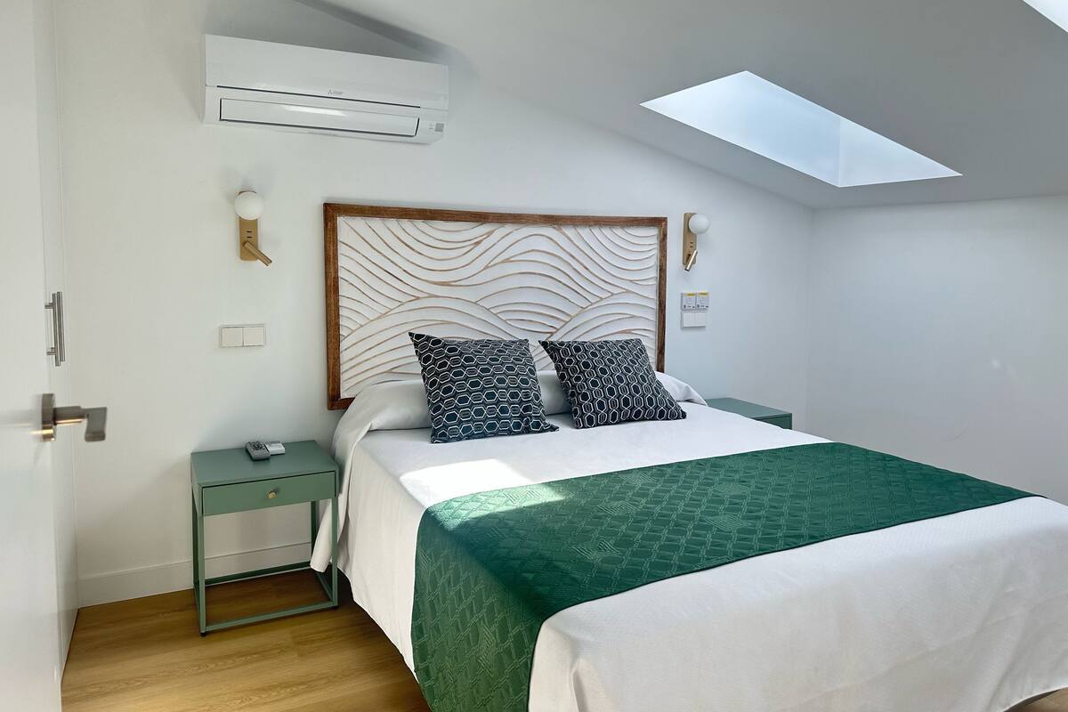 Um quarto moderno com um claraboia, cabeceira de madeira, colcha de cama estampada em verde e uma mesa de cabeceira com um telefone.