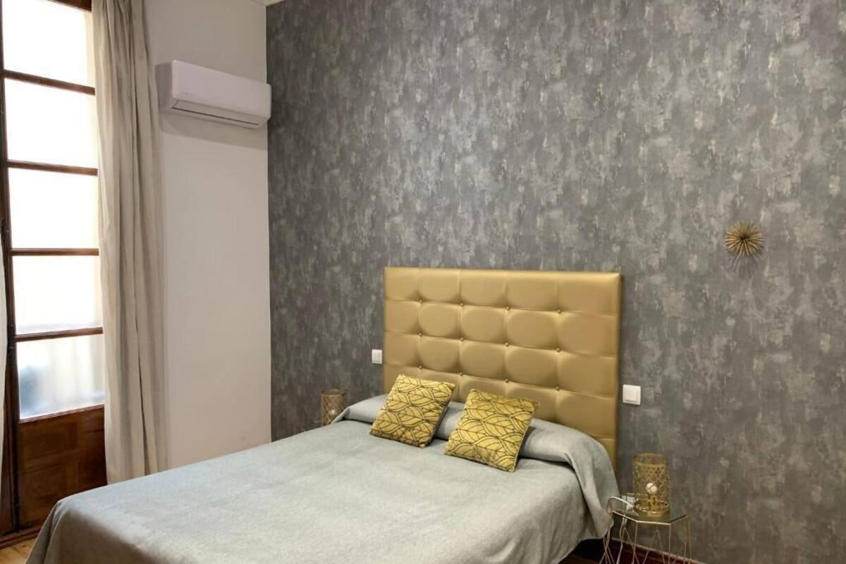 Quarto com cabeceira estofada, duas almofadas, mesa de cabeceira com abajur e janela com cortinas.