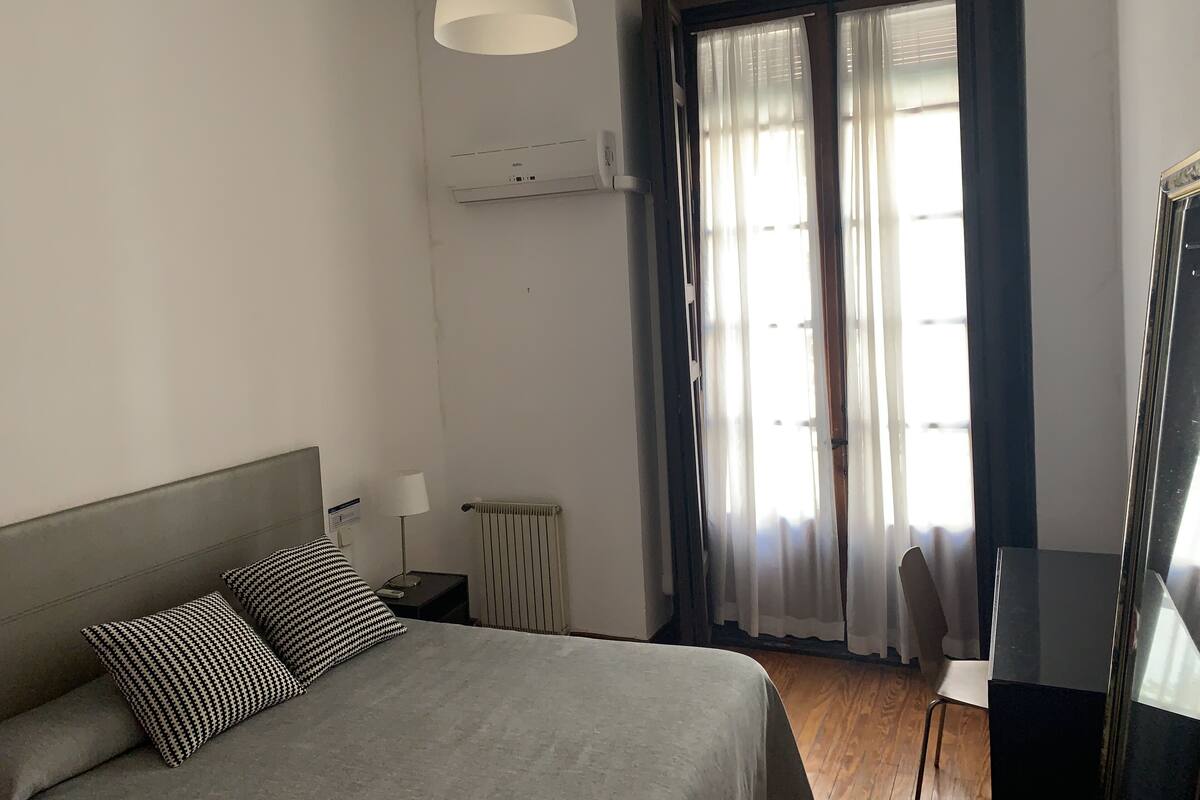 Um quarto com cama, mesa de cabeceira, abajur, radiador e janela com cortinas.