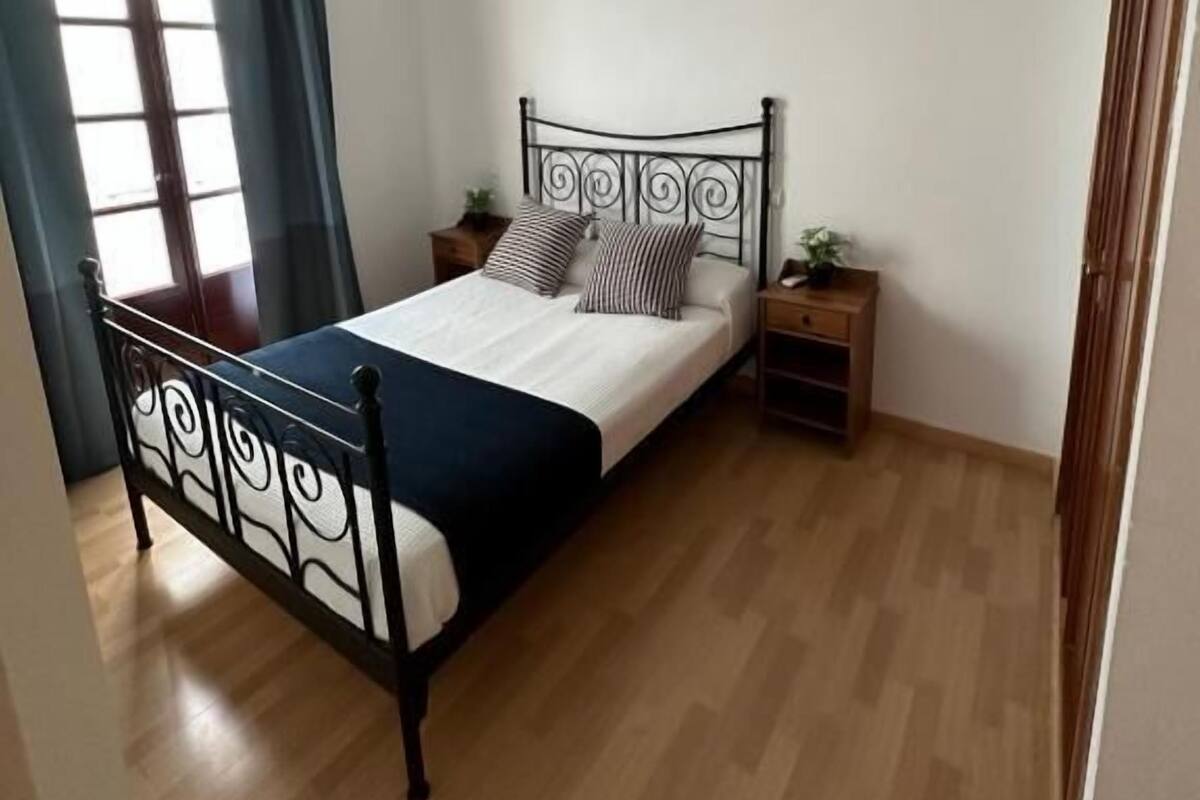 Quarto com cama, mesa de cabeceira, luminária pendente e janela com cortinas.