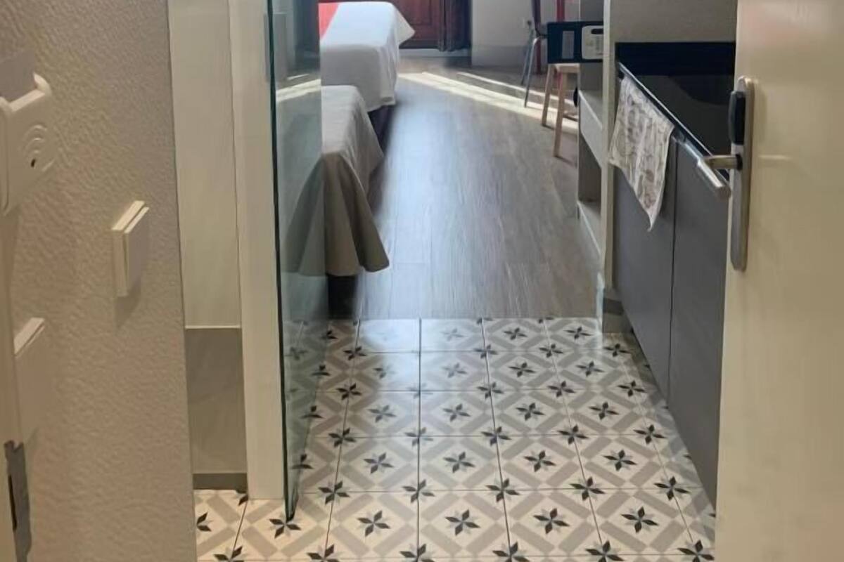 Um corredor estreito que dá para um quarto com uma cama, uma mesa pequena e um refrigerador.