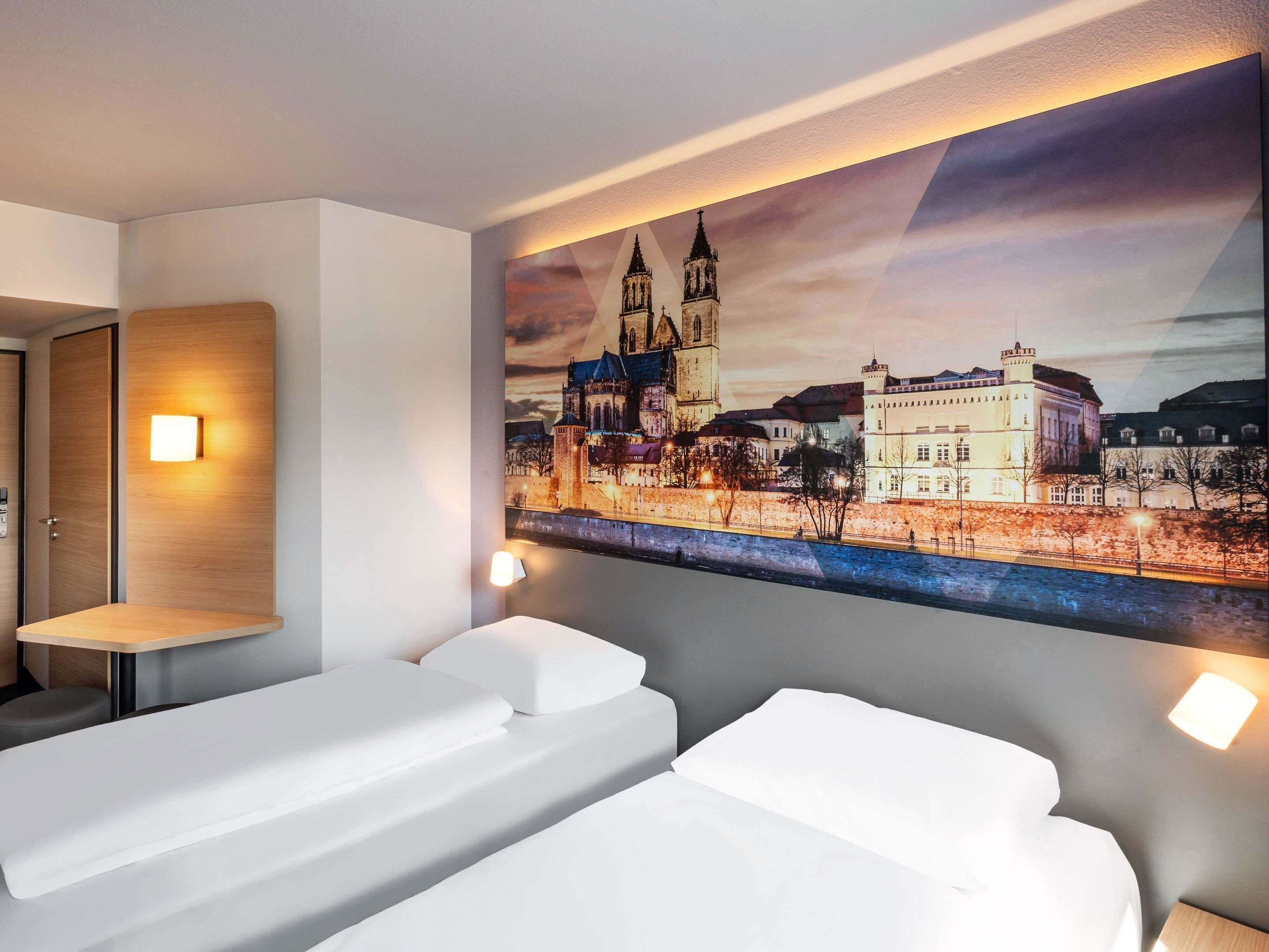 Quarto de hotel com duas camas, uma pintura de paisagem urbana e uma pequena mesa.