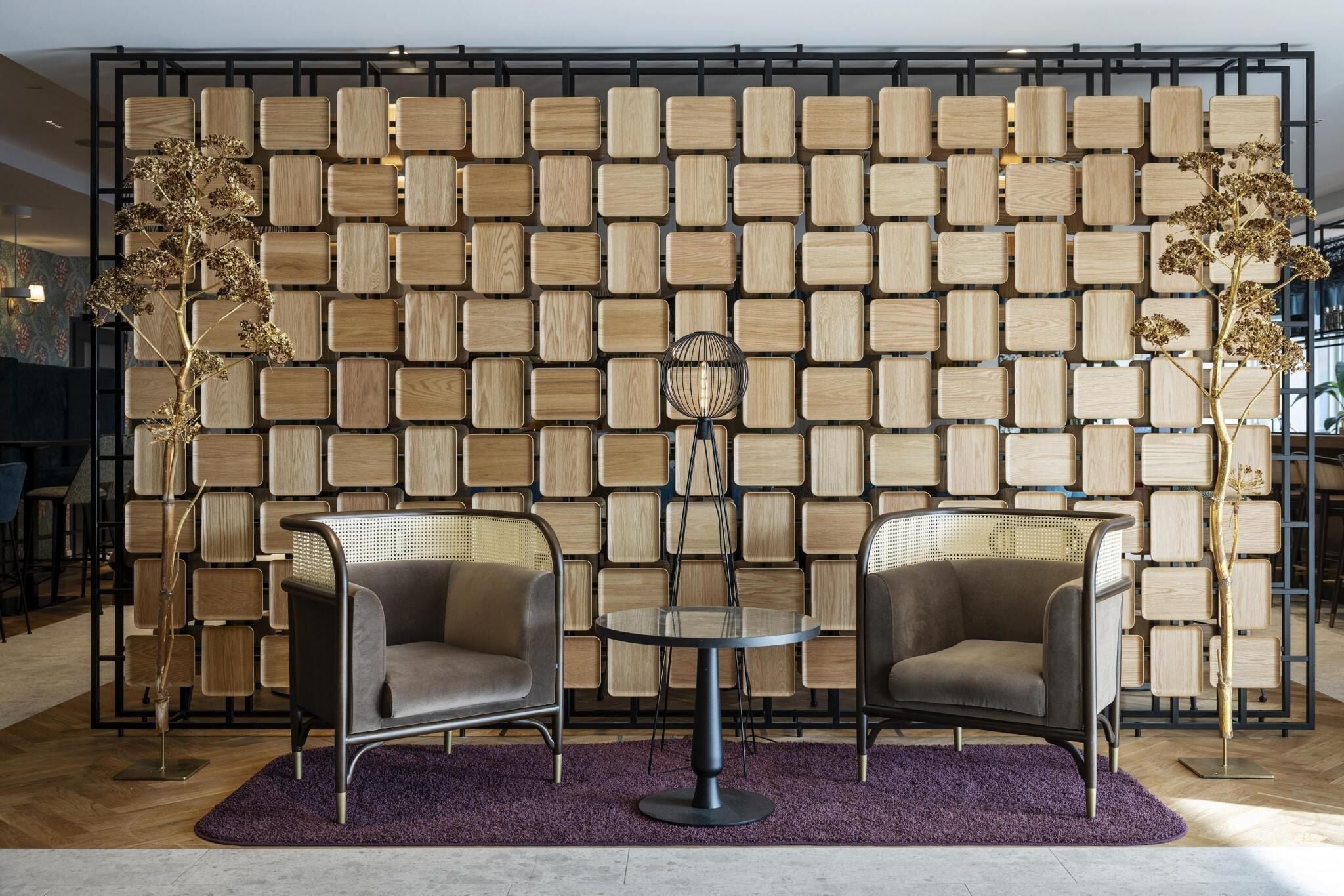 Um interior moderno com parede de painéis de madeira, duas poltronas estofadas, uma mesa redonda e plantas decorativas.
