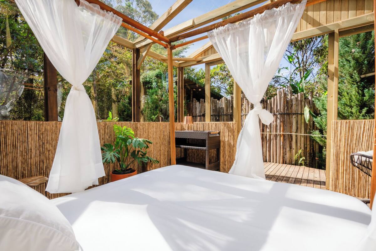 Quarto com cama de dossel, deck de madeira e vegetação ao redor.