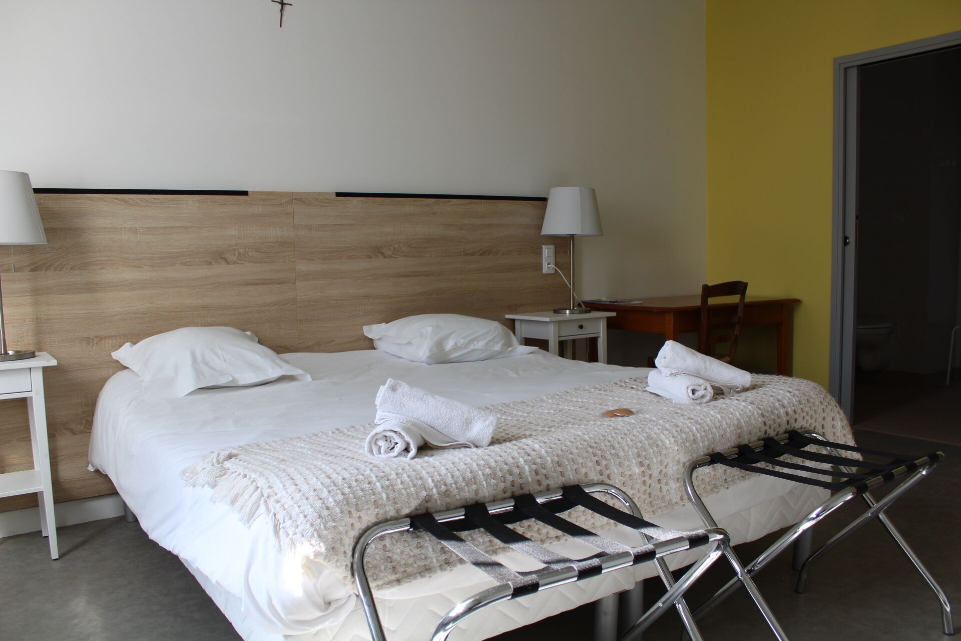 Cama bem arrumada com lençóis e toalhas brancos, cabeceira de madeira, mesa de cabeceira com abajur e uma cadeira em um quarto com parede amarela.