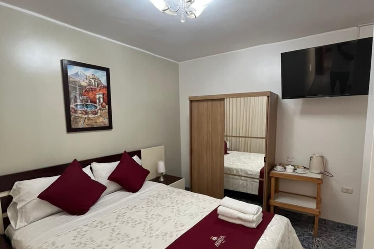 Quarto de hotel com uma cama, televisão, guarda-roupa de madeira, uma pequena mesa com chávenas e uma chaleira, e um quadro na parede.