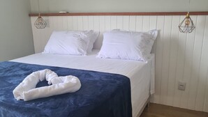 Cama bem arrumada com lençóis brancos e colcha azul, uma toalha branca dobrada sobre a cama e cabeceira de madeira.