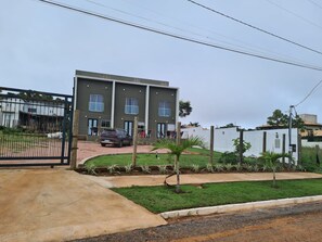 Um edifício moderno de dois andares, com portão fechado, entrada com calçada e pequena área ajardinada com palmeiras.