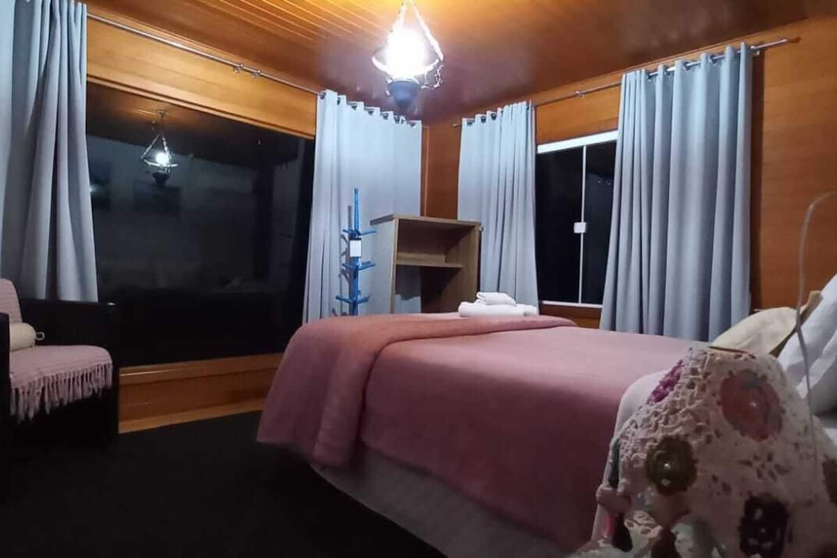 Quarto de hotel com cama, uma cadeira, uma mesa pequena e janela com cortinas.
