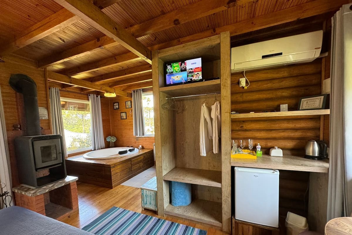 Interior acolhedor de uma cabana, com lareira a lenha, televisão, mini-geladeira e banheira.