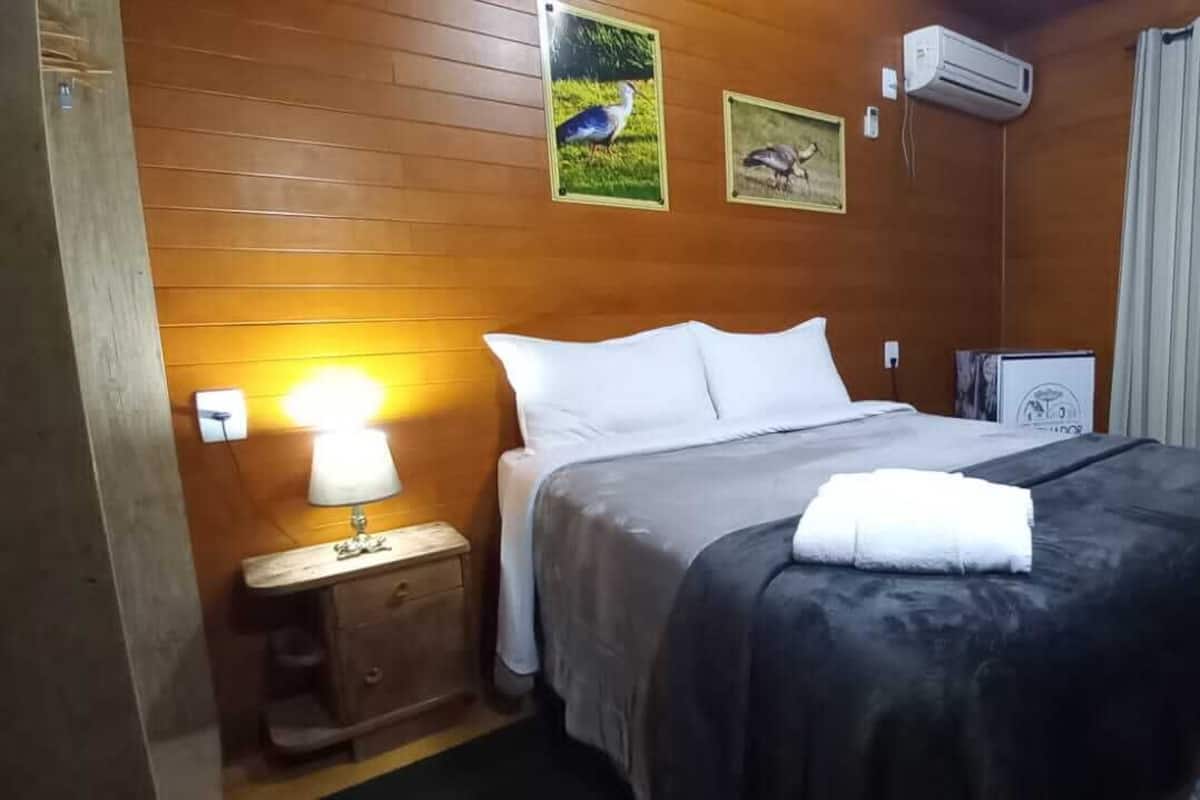 Quarto de hotel com uma cama, uma mesa de cabeceira com abajur, uma cadeira vermelha e duas imagens emolduradas na parede.