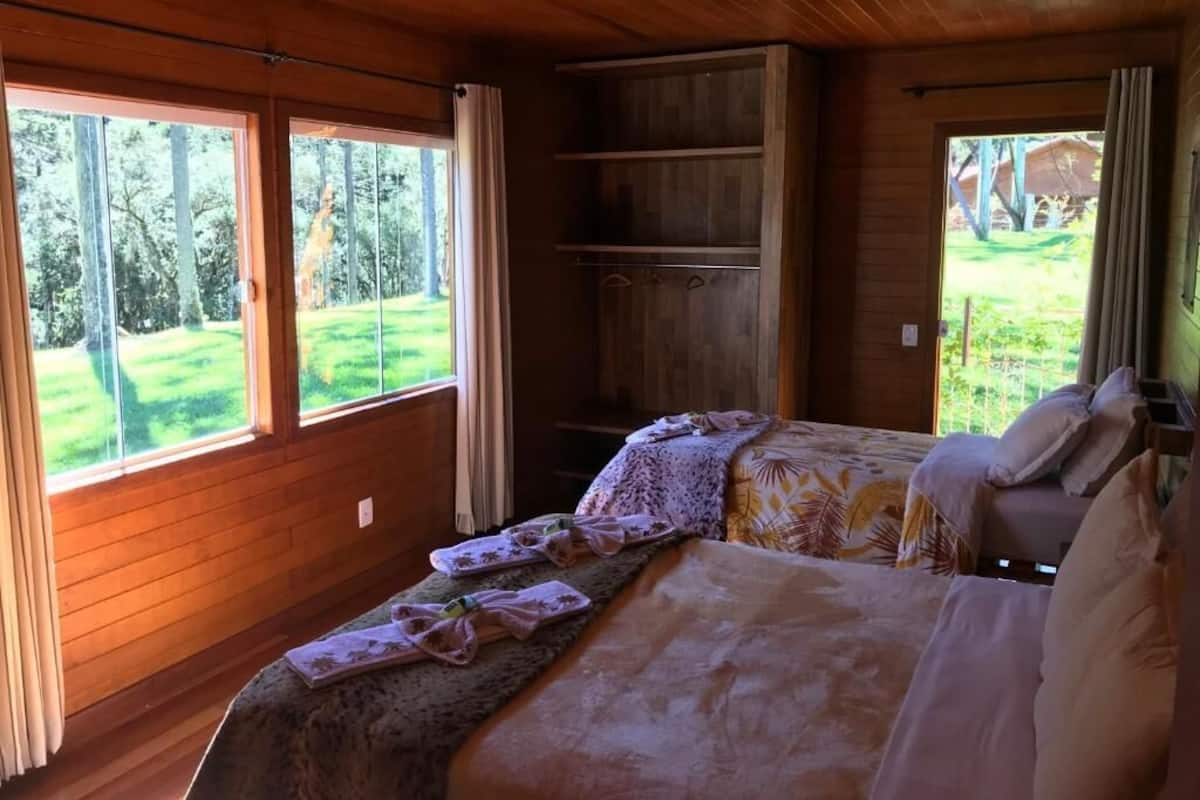 Quarto de cabana de madeira com duas camas, um guarda-roupa embutido e janelas amplas com vista para uma paisagem gramada.