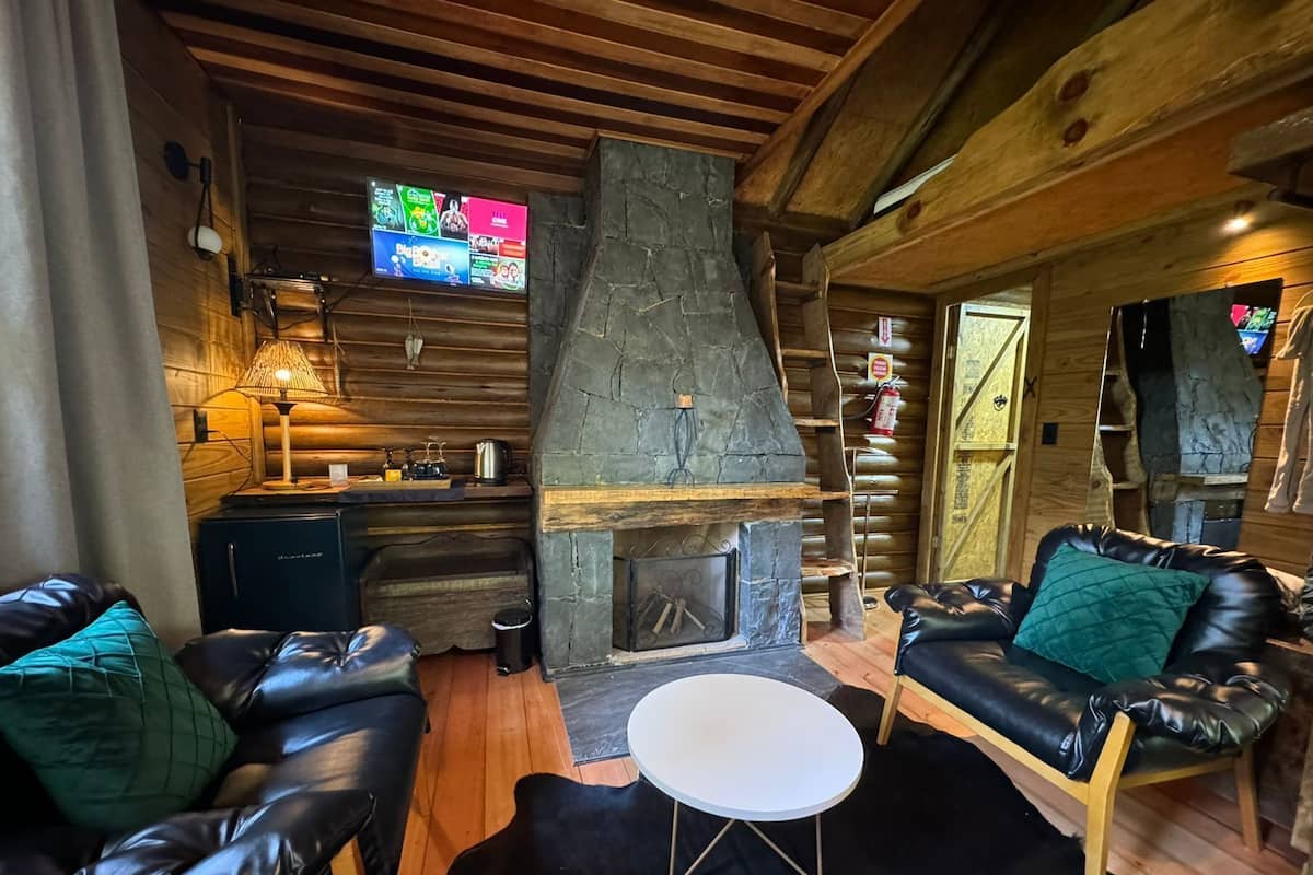 Interior acolhedor de uma cabana de madeira com lareira de pedra, sofás de couro, uma mesa redonda pequena e uma televisão fixada na parede.