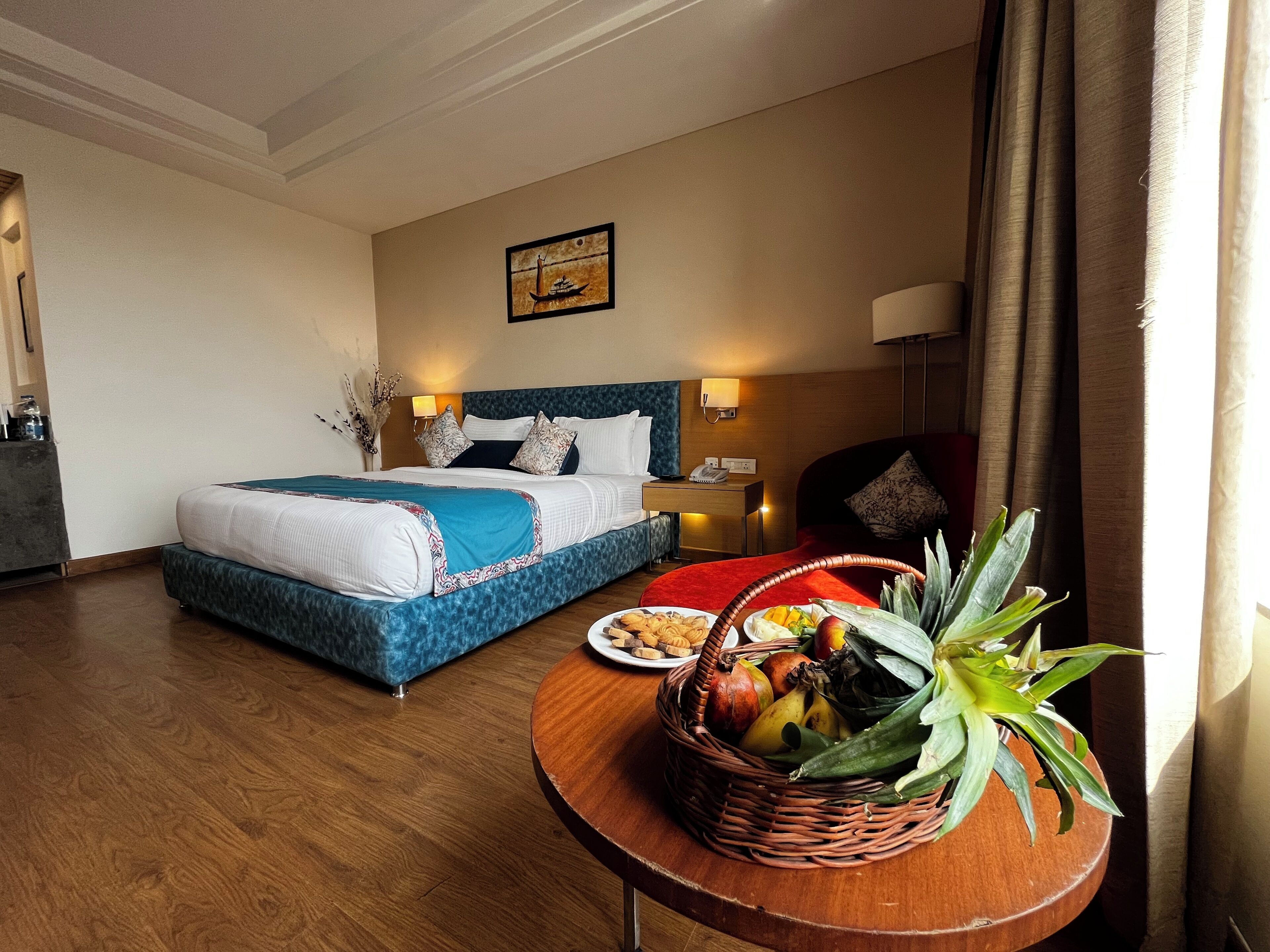 Quarto de hotel com cama, mesa de cabeceira, abajur e uma cesta de frutas sobre uma mesa.