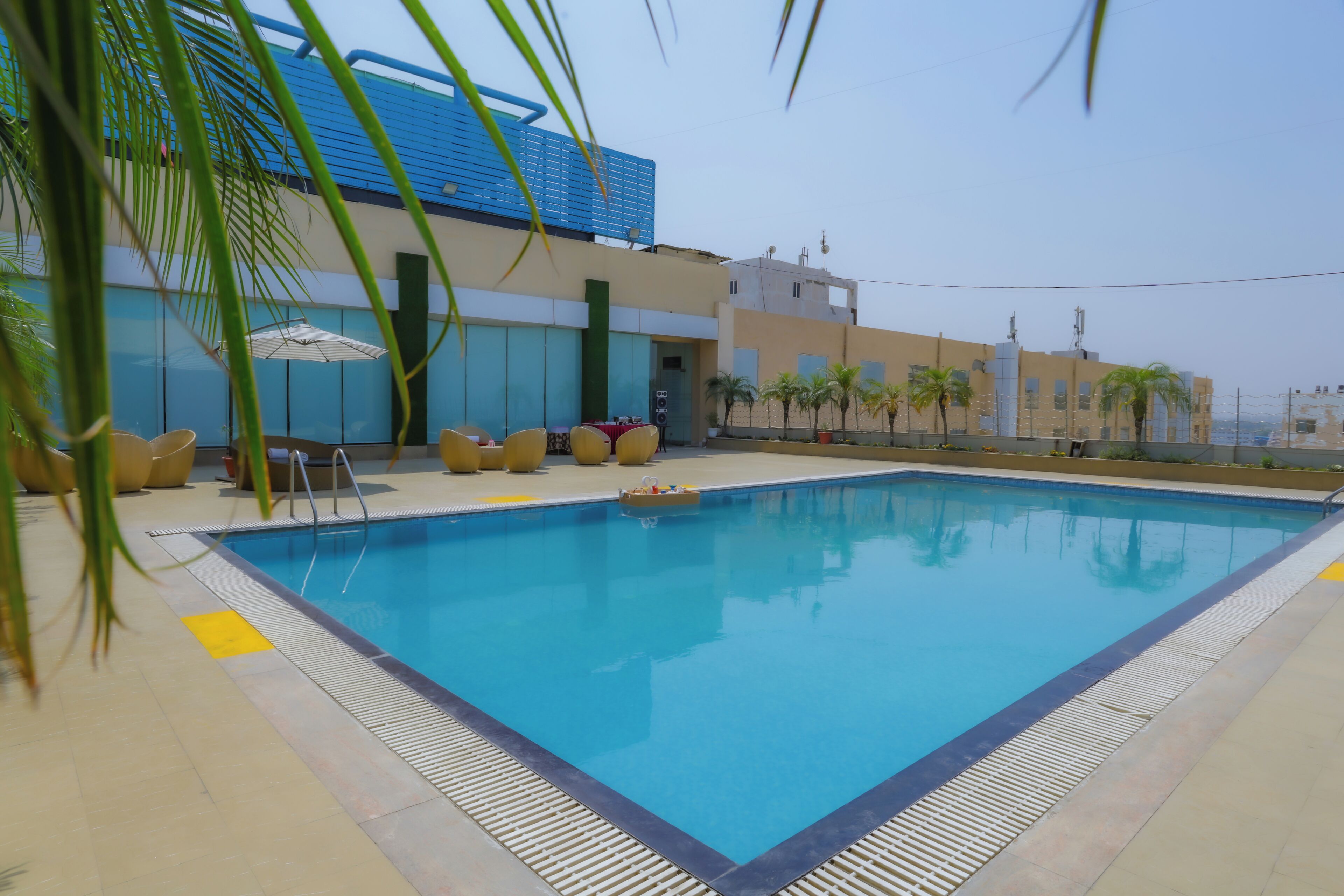 Piscina com cadeiras de descanso e guarda-sóis, cercada por edifícios e palmeiras.