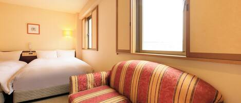Quarto de hotel com duas camas, um sofá listrado, uma mesa de cabeceira com abajur e uma janela com vista.