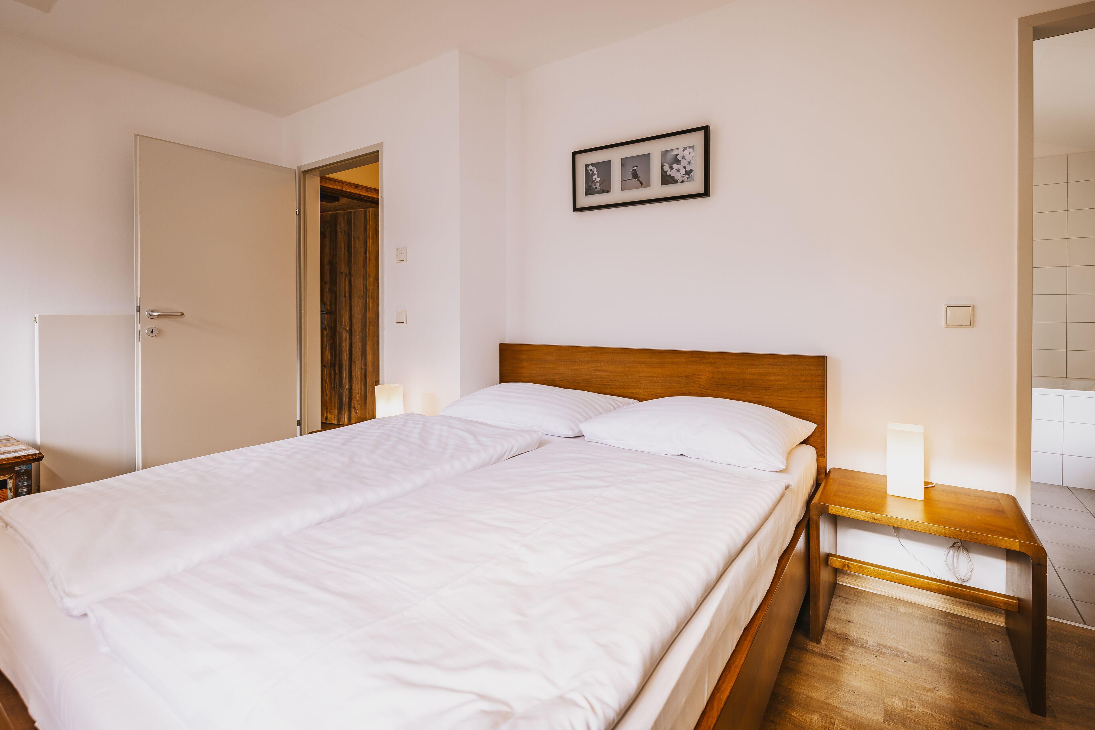 Cama bem arrumada com lençóis brancos, cabeceira de madeira e mesa de cabeceira com abajur.