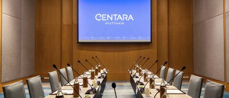 Uma sala de conferências com uma mesa comprida, cadeiras, microfones e uma tela exibindo 'CENTARA AYUTTHAYA'.