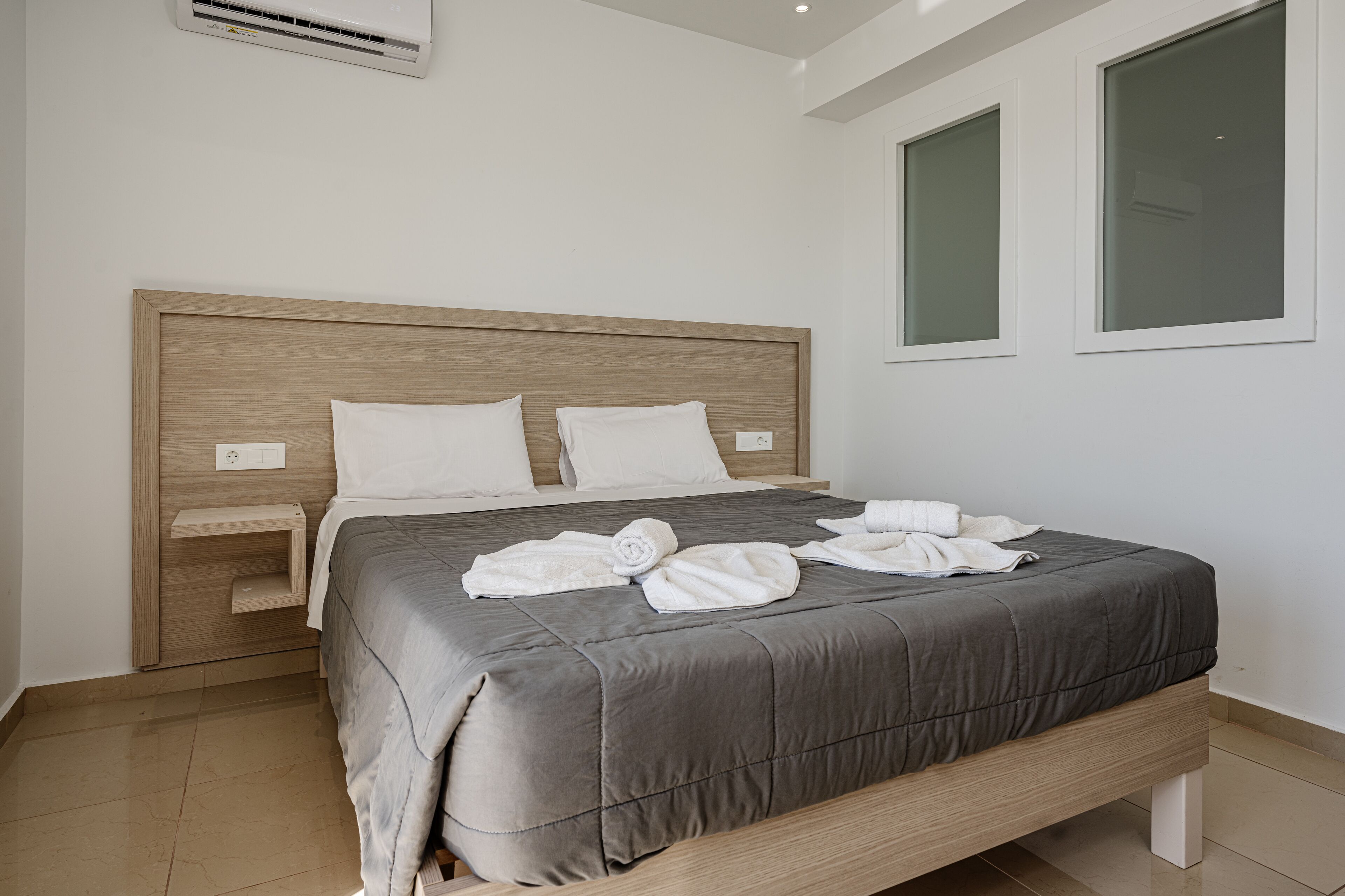 Quarto moderno com cabeceira de madeira, colcha cinza e toalhas brancas.