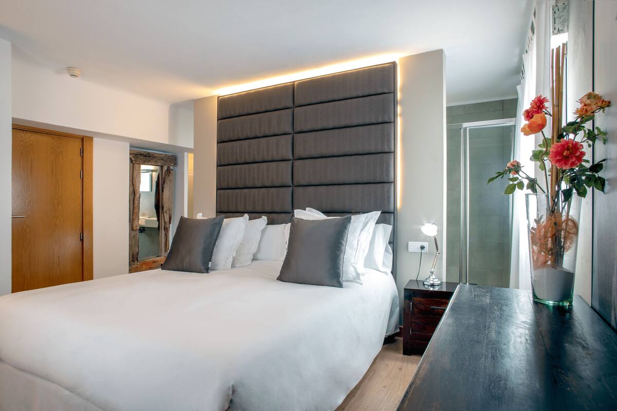 Quarto de hotel moderno com uma cama grande, um guarda-roupa de madeira, uma mesa de cabeceira com abajur e um vaso com flores.