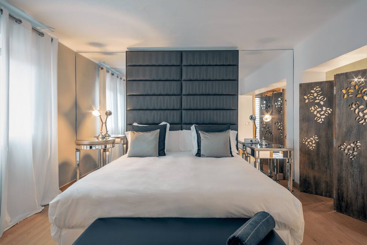 Quarto de hotel moderno com uma cama grande, mesas de cabeceira, um espelho e um painel decorativo.