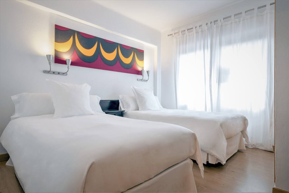 Quarto de hotel com duas camas, um quadro moderno na parede e uma janela grande com cortinas leves.
