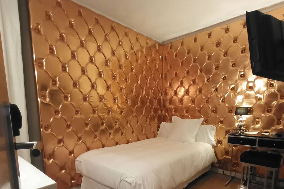 Quarto com cabeceira estofada, cama com roupa de cama branca, mesa de cabeceira e escrivaninha com cadeira.