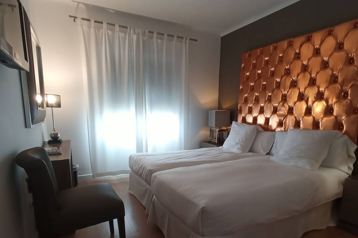 Quarto de hotel com uma cama grande, uma cadeira, uma mesa e uma janela com cortinas.