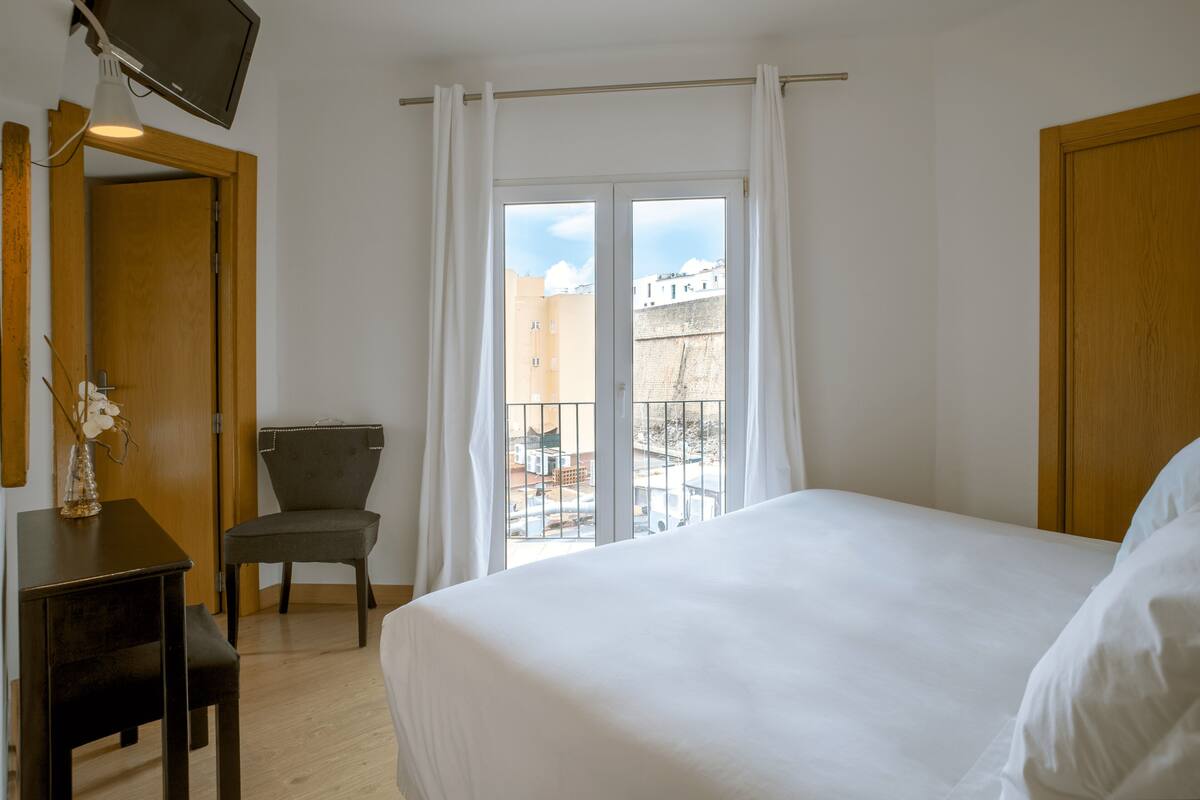 Quarto de hotel com cama, escrivaninha, cadeira, televisão e varanda com vista para edifícios.