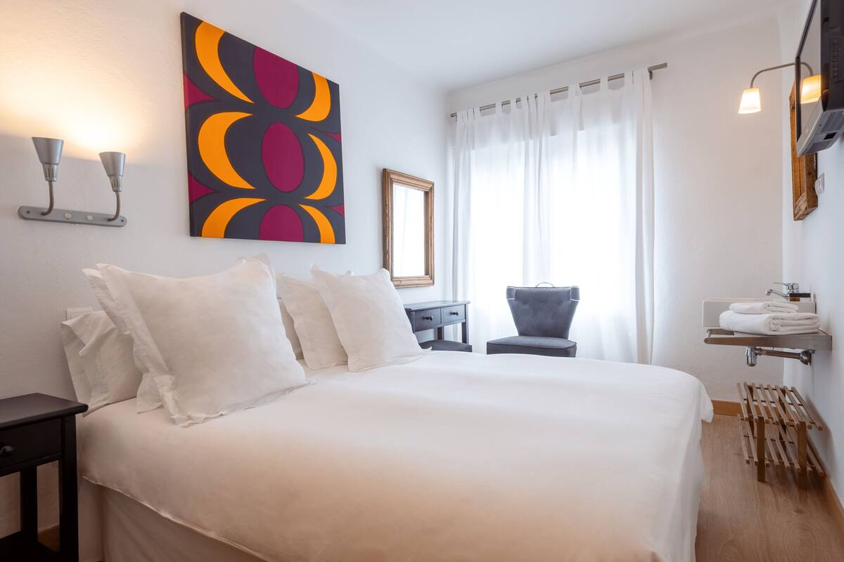 Quarto de hotel com uma cama grande, uma escrivaninha, uma cadeira, banheiro com porta-toalhas e um quadro abstrato colorido na parede.