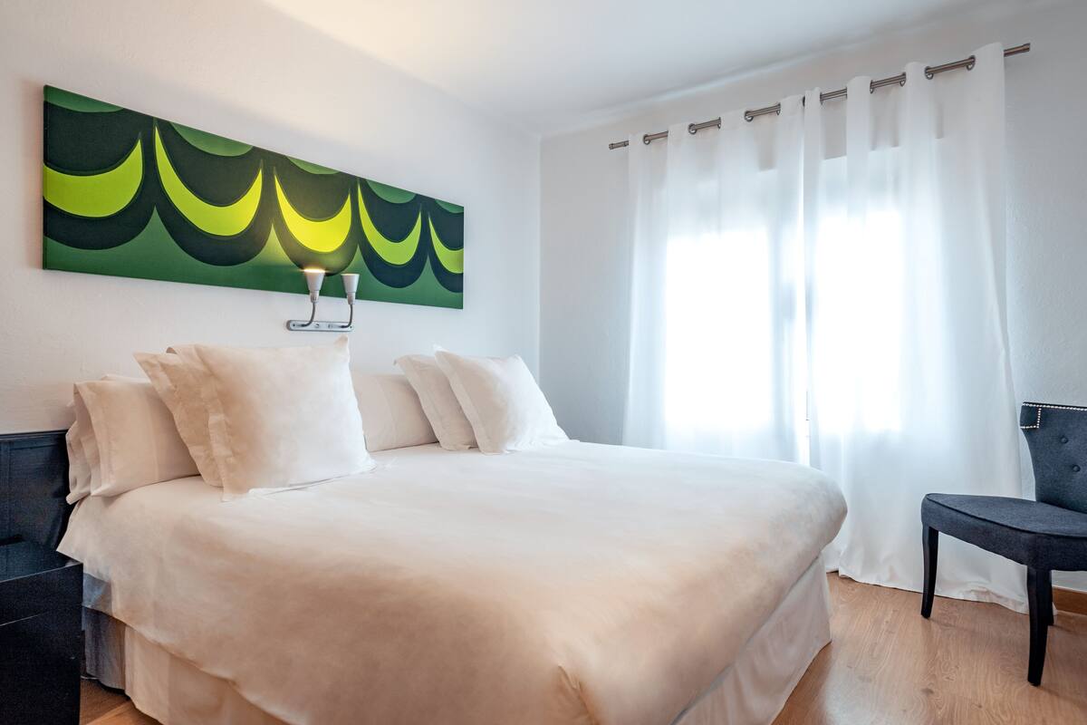 Uma cama bem arrumada com lençóis brancos, uma pintura abstrata moderna na parede e uma cadeira junto à janela.