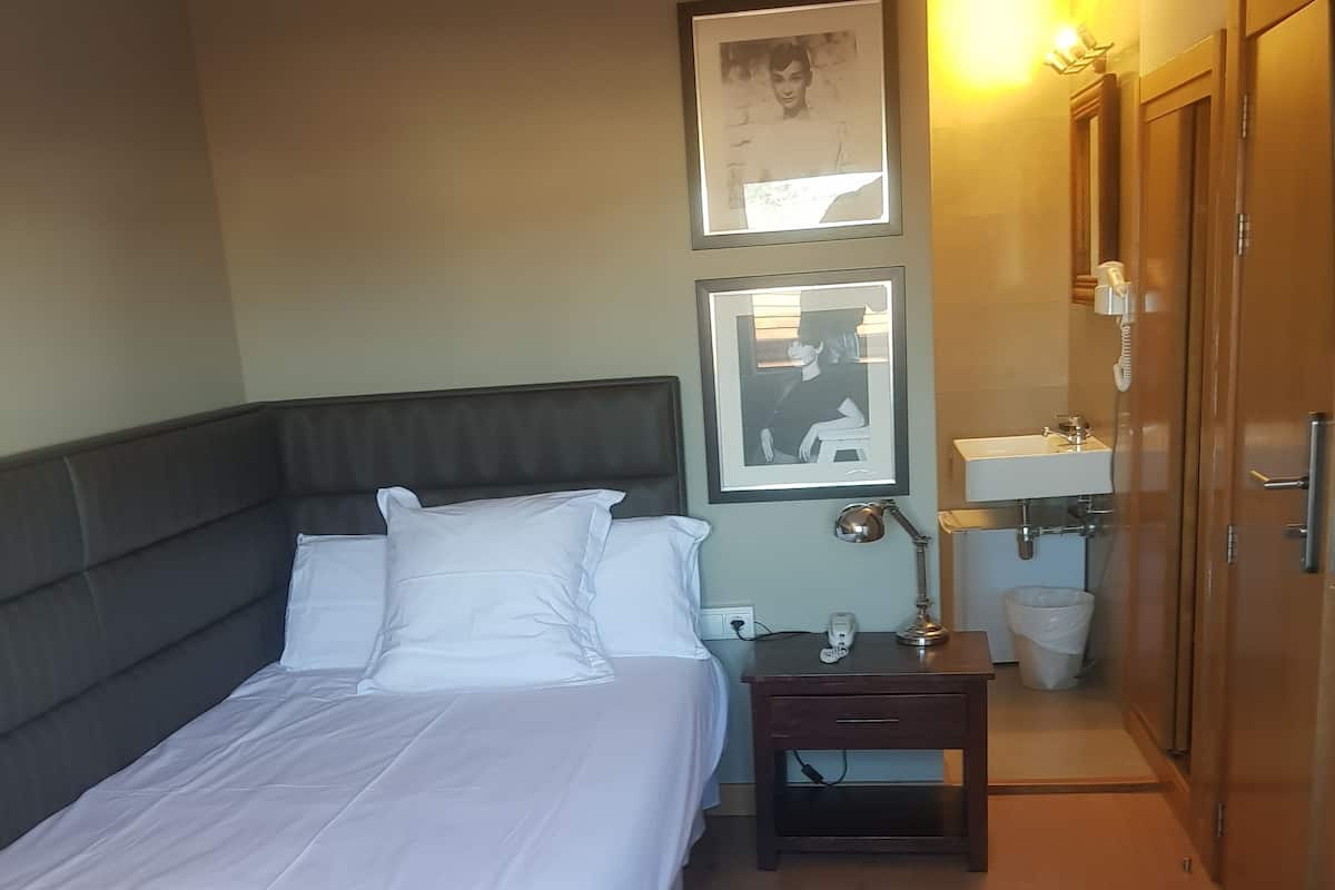 Quarto de hotel com uma cama grande, mesa de cabeceira com abajur, quadro na parede, banheiro pequeno com pia e um guarda-roupa.
