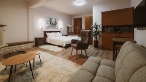 Uma sala de estar moderna com um sofá, uma poltrona, uma mesinha e uma cama com colcha branca. Há uma lareira, um quadro na parede e uma área de cozinha ao fundo.