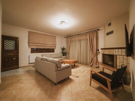 Uma sala de estar com um sofá bege, uma mesa de centro de madeira, uma lareira e uma TV fixada na parede.