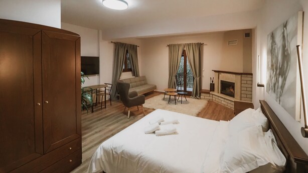 Quarto de hotel com uma cama grande, um guarda-roupa de madeira, uma pequena mesa com cadeiras, um sofá, uma lareira e uma janela com cortinas.