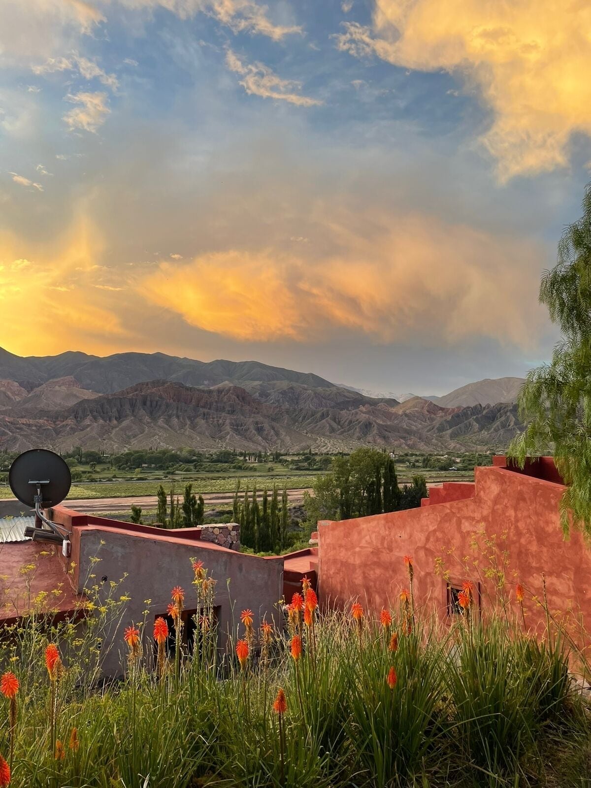 Paisagem montanhosa com pôr do sol, apresentando um edifício com antena parabólica e paredes vermelhas, cercado por vegetação verde e flores laranja.