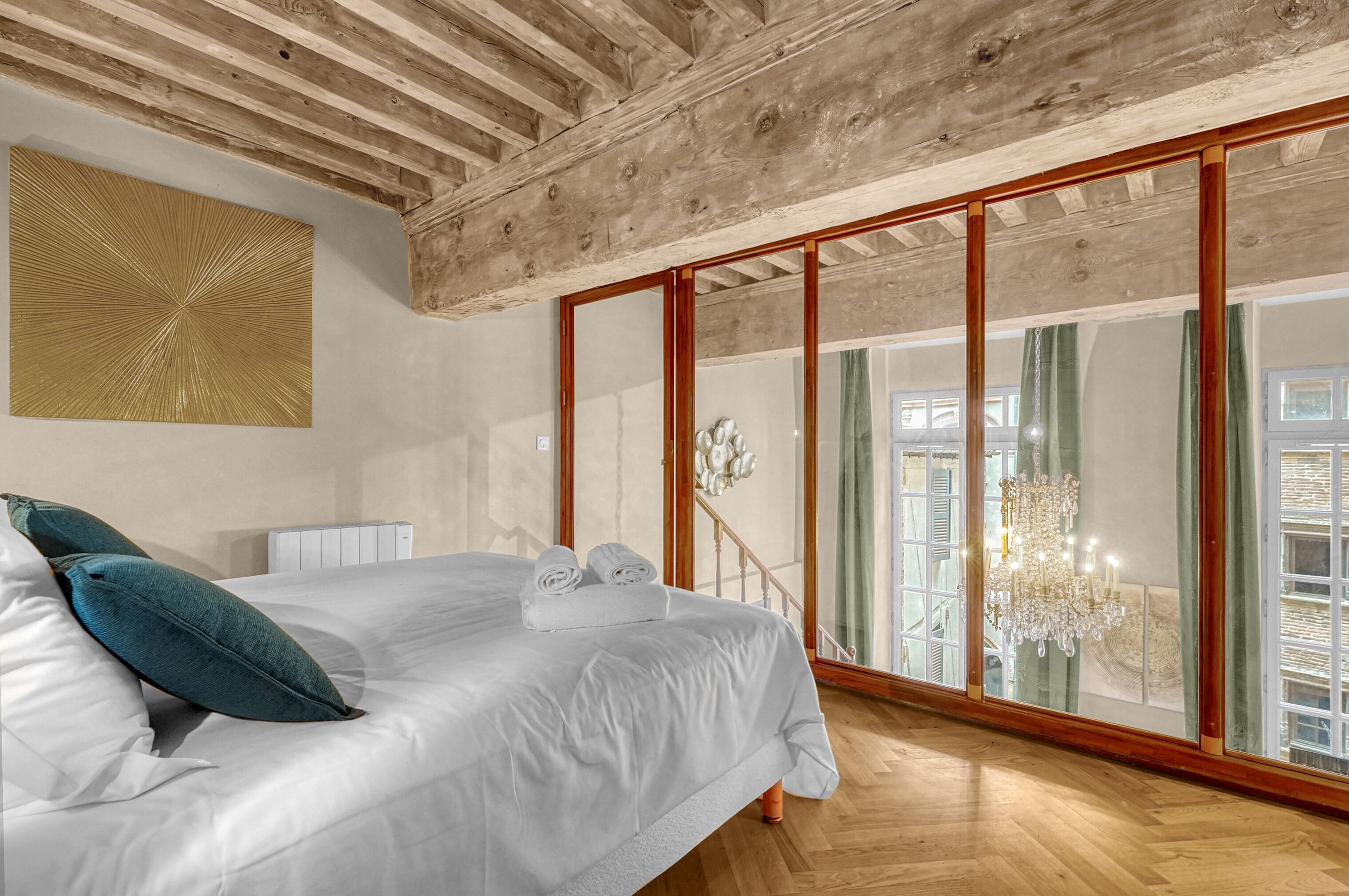 Quarto com cama grande, vigas de madeira no teto, vista por uma porta de vidro e um lustre.