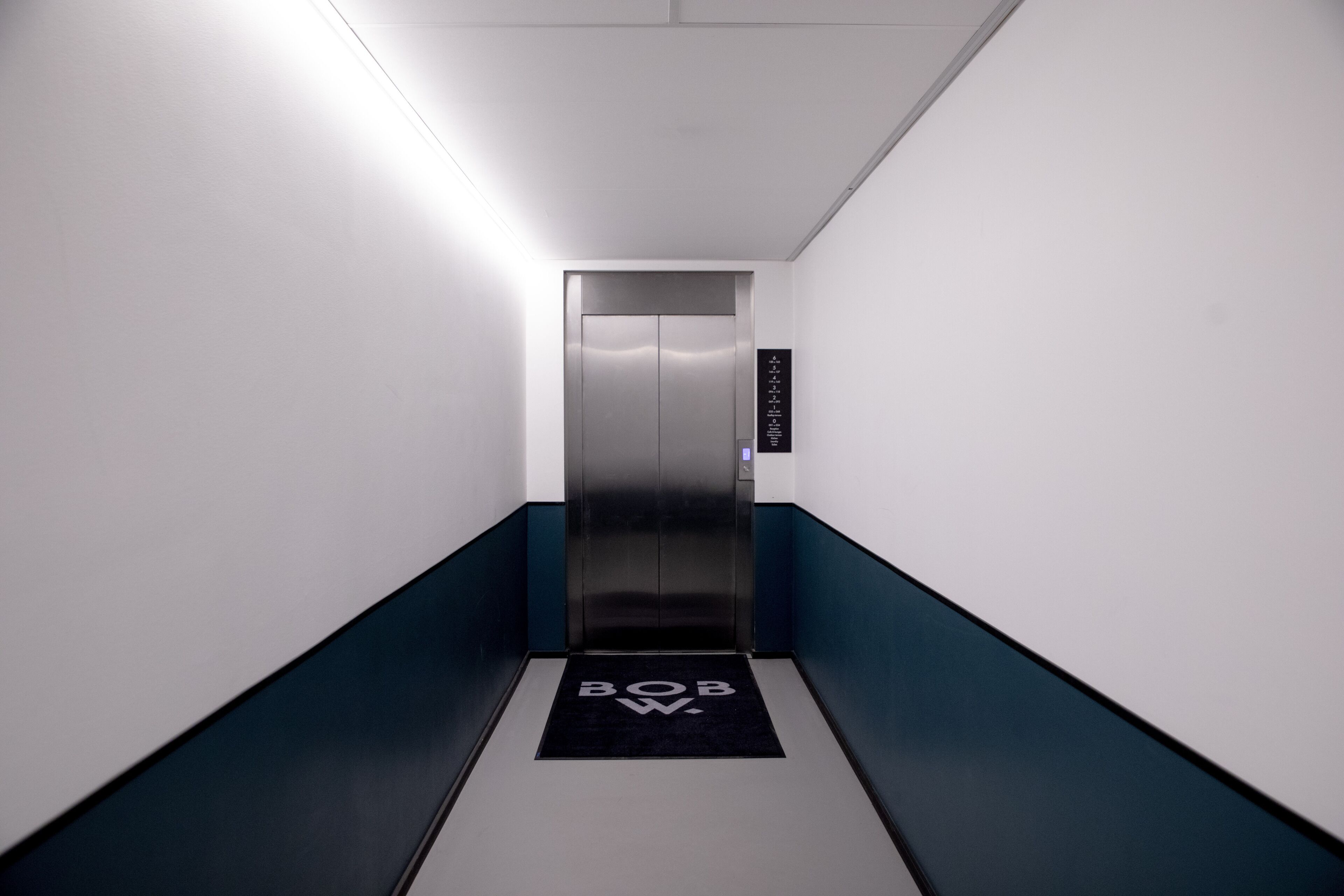 Um corredor estreito com tapete preto e branco com a inscrição "BOB W." que leva a um elevador metálico com painel de controle.