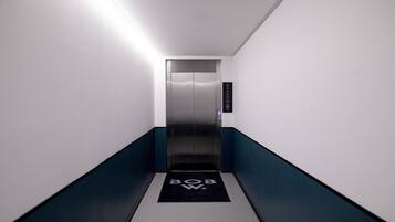 Um corredor estreito com tapete preto e branco com a inscrição "BOB W." que leva a um elevador metálico com painel de controle.