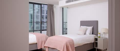 Quarto de hotel moderno com duas camas, uma pequena mesa de cabeceira, uma luminária e vista para a cidade através de grandes janelas.