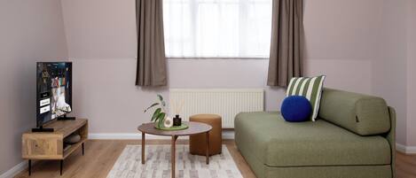 Uma sala de estar com um sofá verde, uma mesa de madeira pequena, um banquinho redondo, uma televisão sobre um suporte de madeira e uma janela com cortinas.