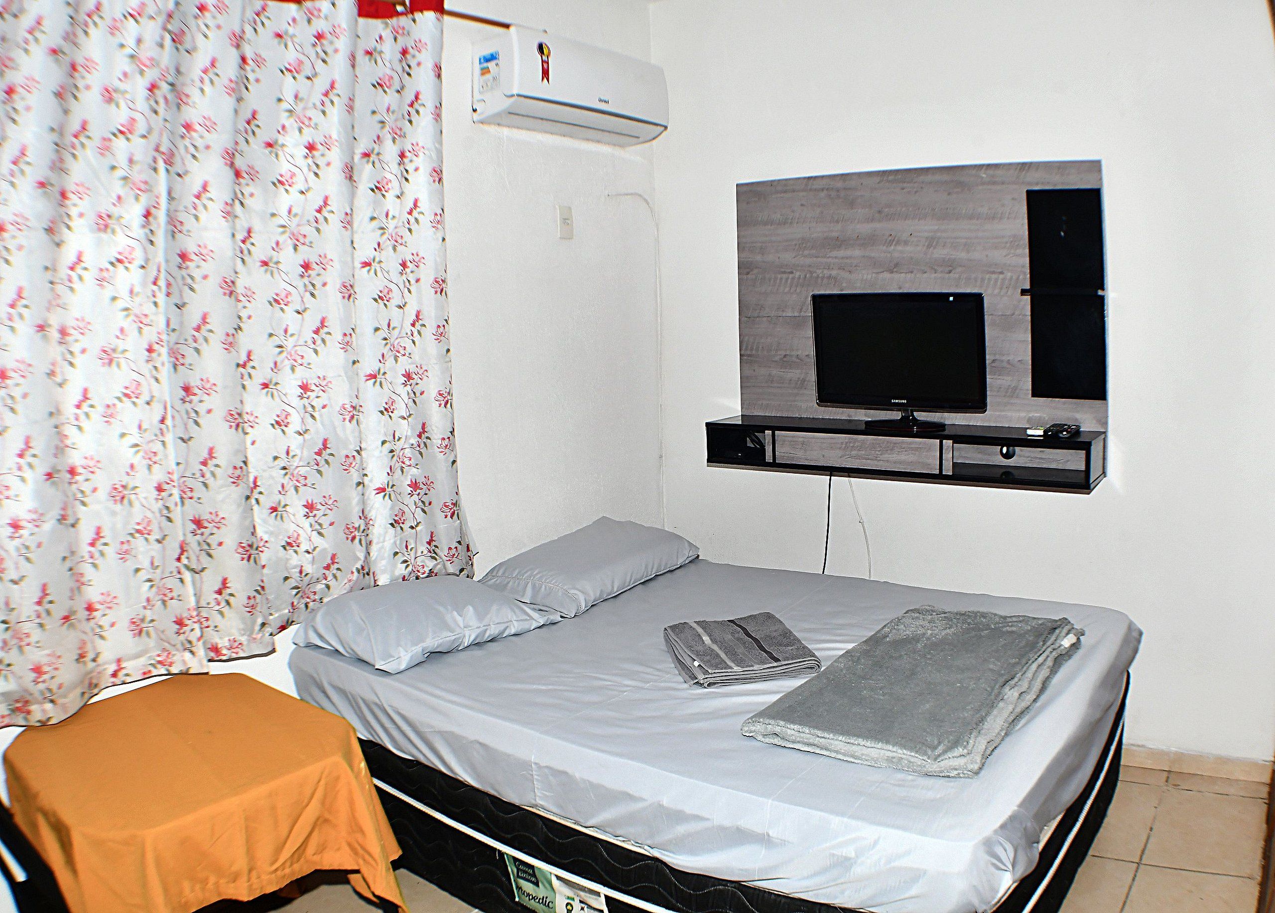 Quarto com cama, televisão na parede, mesa pequena e cortina com padrão floral.