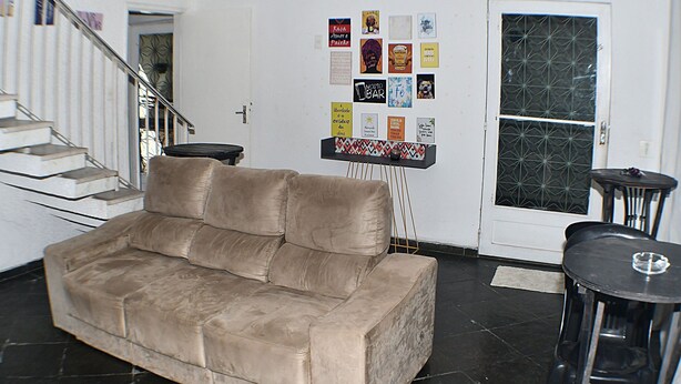 Sala de estar com sofá bege, escada, porta com painel de vidro e parede decorada com vários posters.