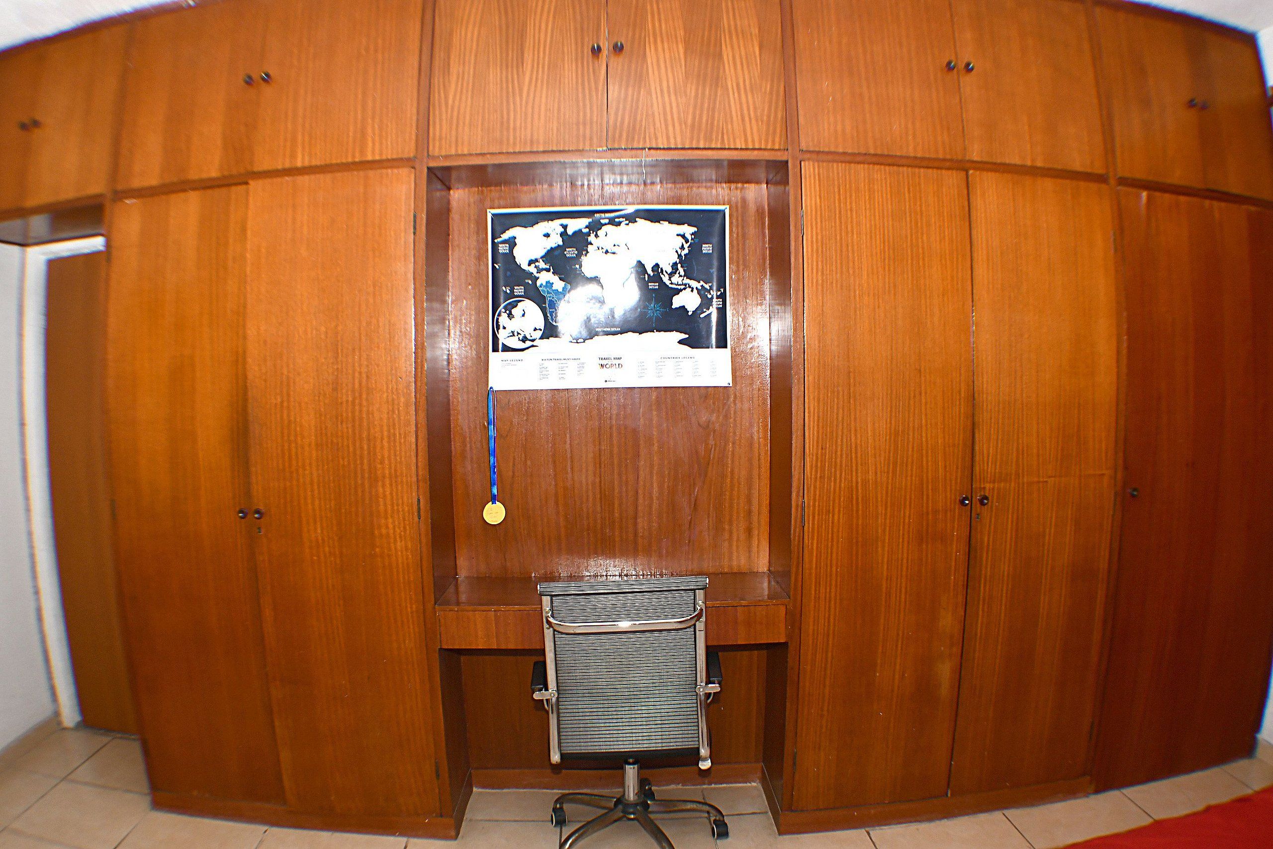 Guarda-roupa de madeira com um cartaz de mapa de viagens, uma pequena escrivaninha com cadeira e um tapete vermelho.