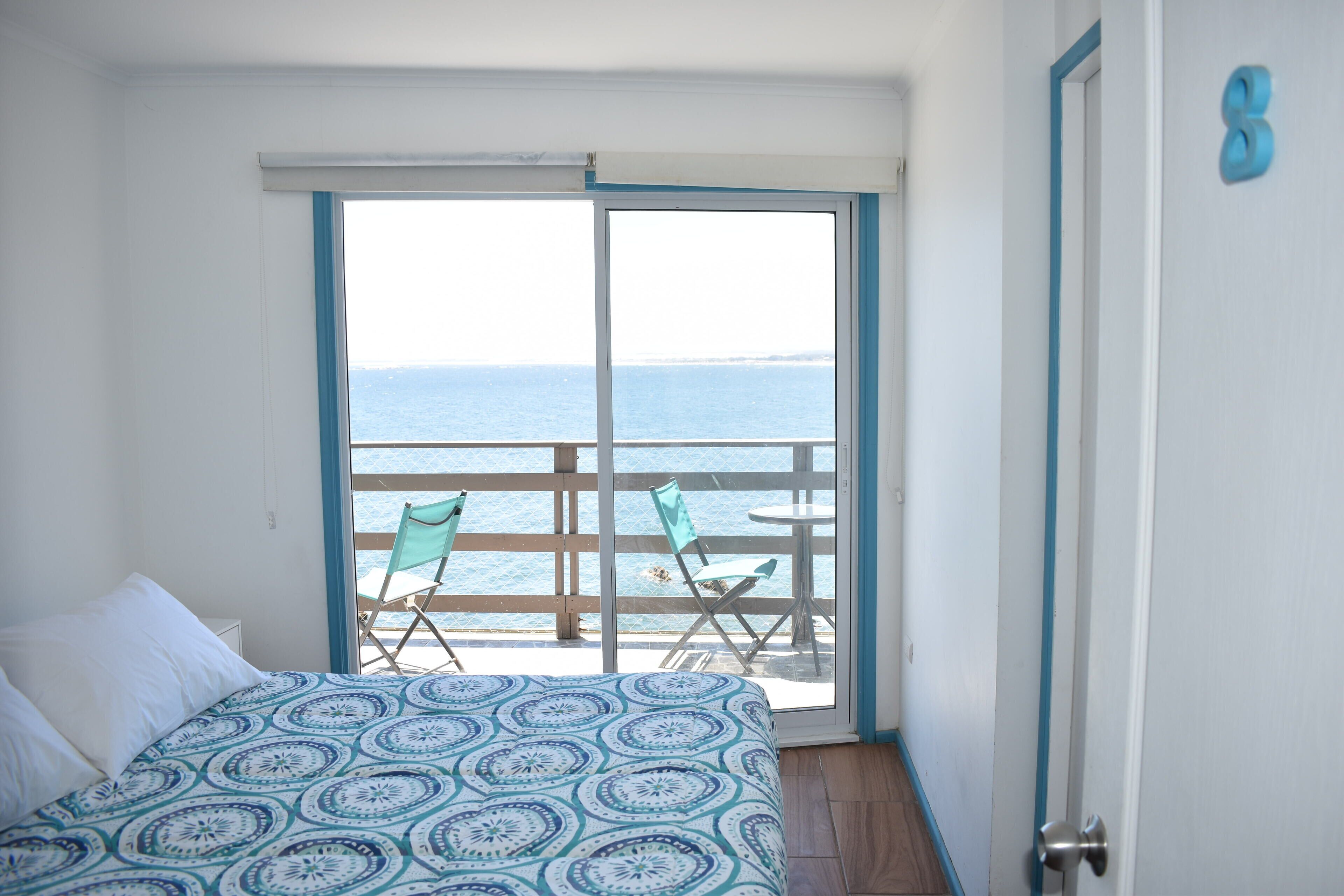 Quarto com cama, varanda com cadeiras e vista para o mar.