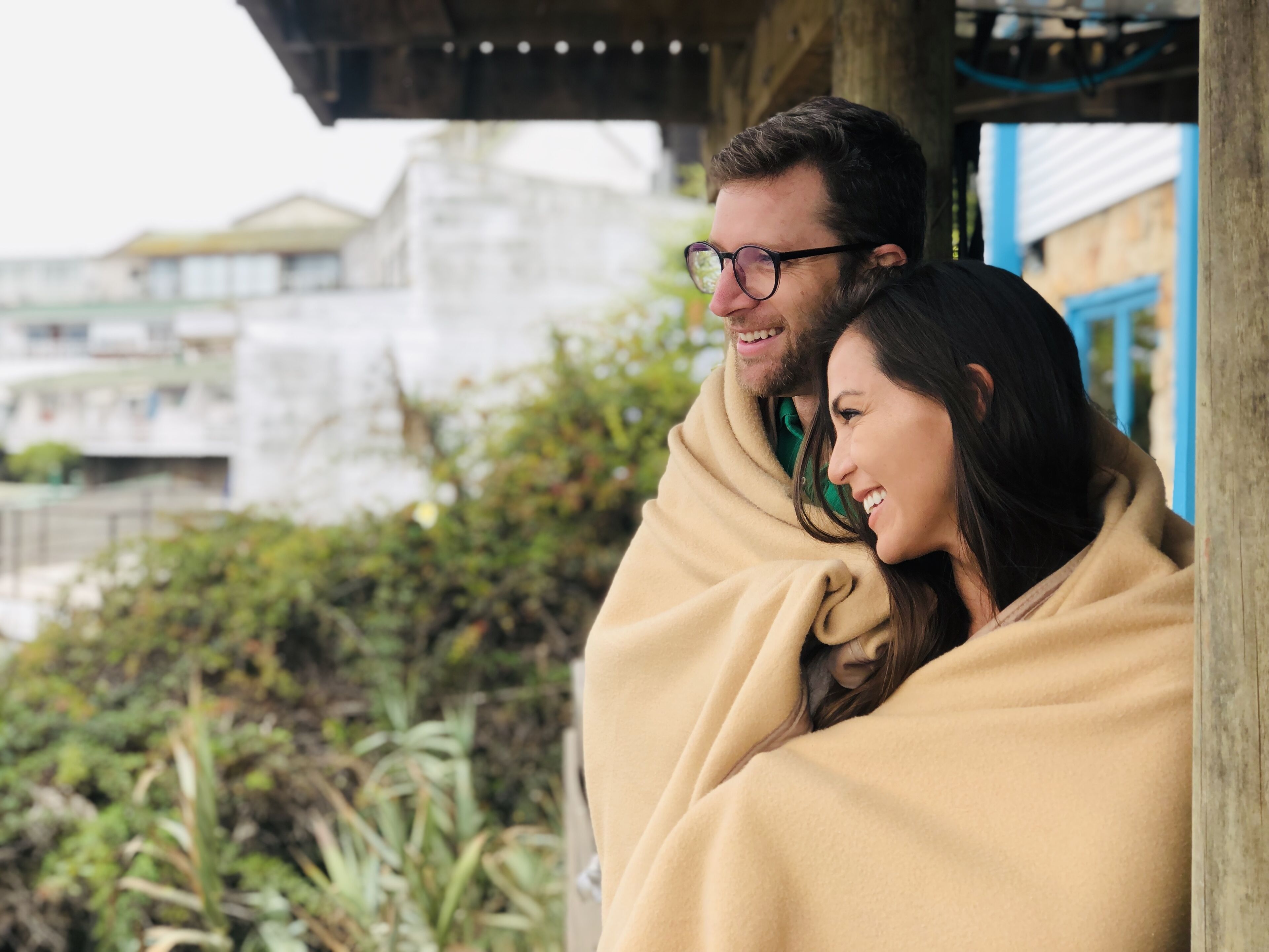 Um casal enrolado em um cobertor, sob uma estrutura de madeira, com vista para edifícios e vegetação ao fundo.